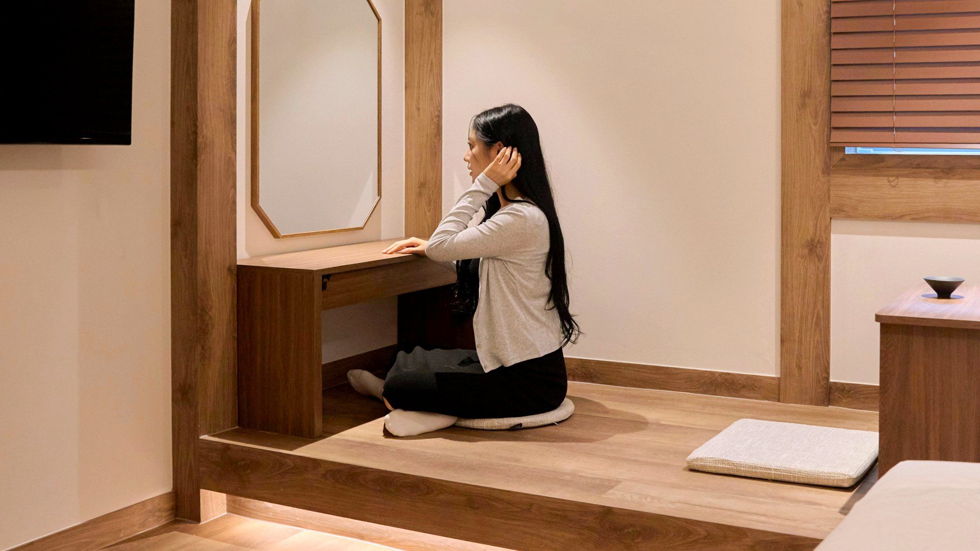 Uma pessoa sentada num banco de madeira em um quarto com piso de madeira, um espelho fixado na parede e uma pequena mesa com uma tigela.