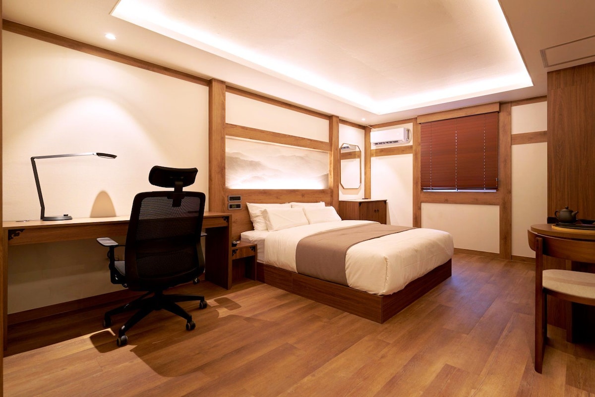 Quarto de hotel com cama, escrivaninha, cadeira e televisão fixada na parede.