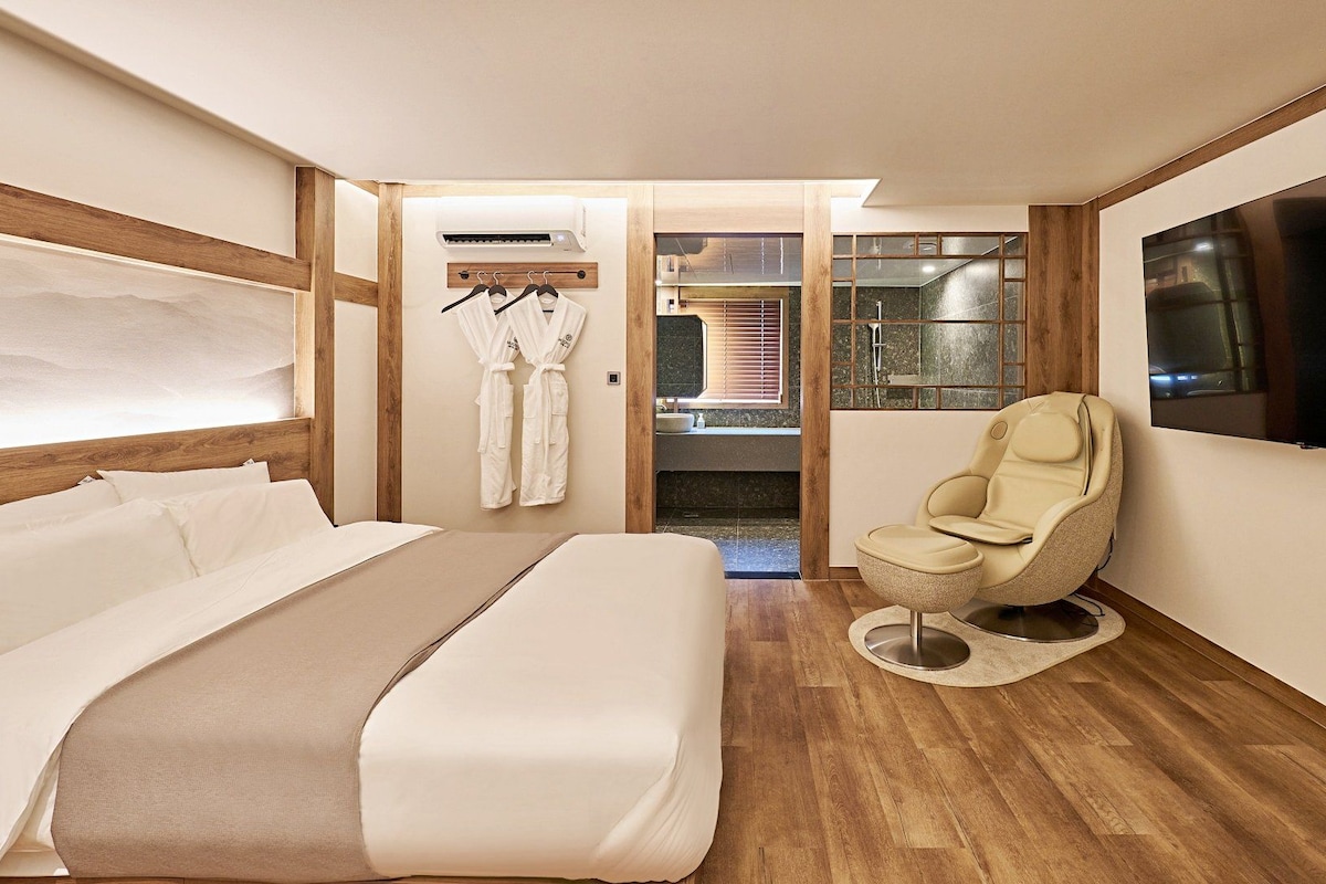 Quarto de hotel moderno com uma cama grande, poltrona de massagem, televisão e vista para o banheiro.