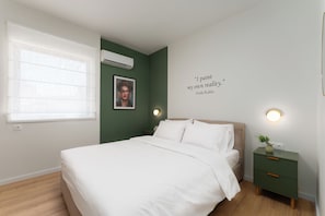 Quarto com cama, mesa de cabeceira, ar-condicionado fixado na parede, quadro emoldurado e frase motivacional.