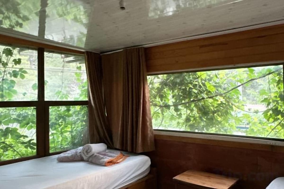 Quarto com uma cama de solteiro, um banco de madeira e uma grande janela com cortinas.