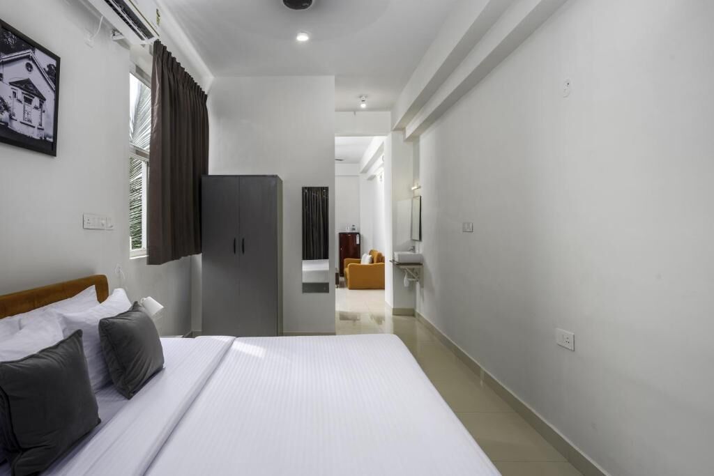 Quarto de hotel com cama, guarda-roupa, banheiro com pia e espelho, e corredor que dá para outro quarto.