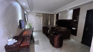 Quarto de hotel com uma mesa de jantar de madeira, um sofá, uma TV fixada na parede e uma porta com puxador.