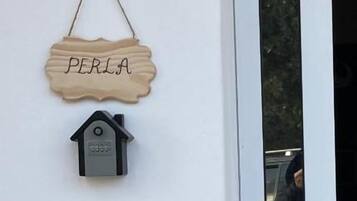 Placa de madeira com o nome 'PERLA' e um nicho de pássaros numa parede, uma porta com painel de vidro e uma parede de pedra.