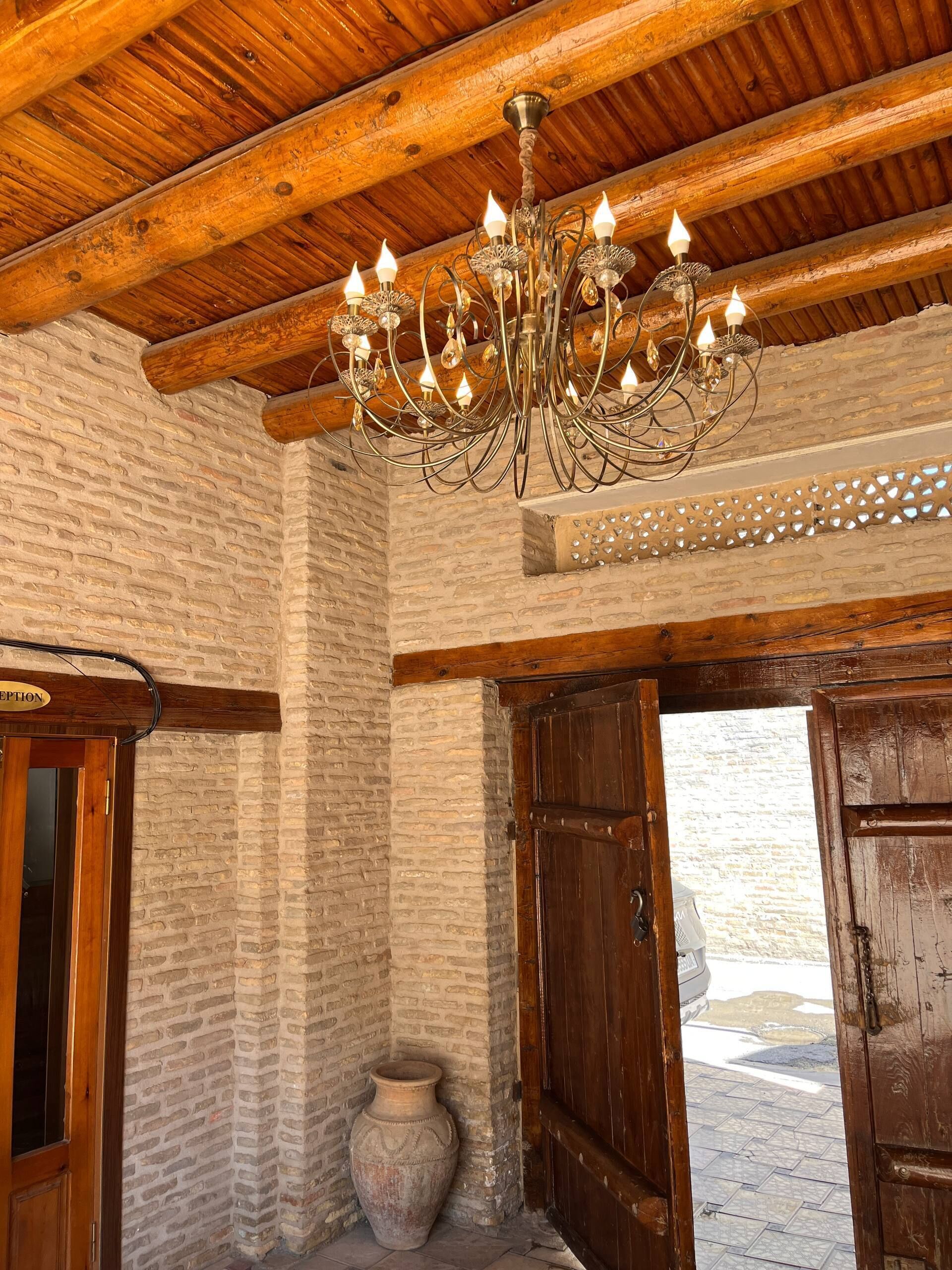 Quarto com teto de madeira, parede de tijolos e um candelabro. Há uma porta de madeira e um vaso grande no chão.