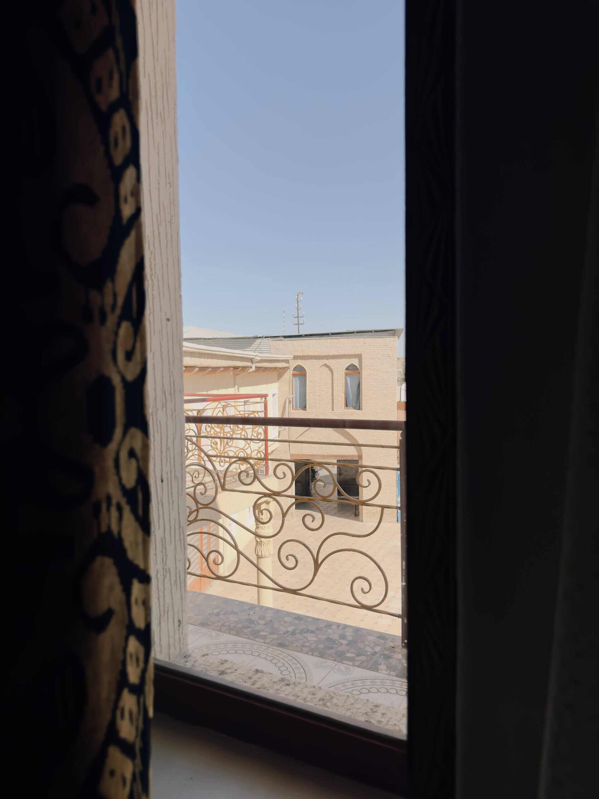 Janela com vista para um edifício com telhado em telhas e varanda com grades de metal decoradas.