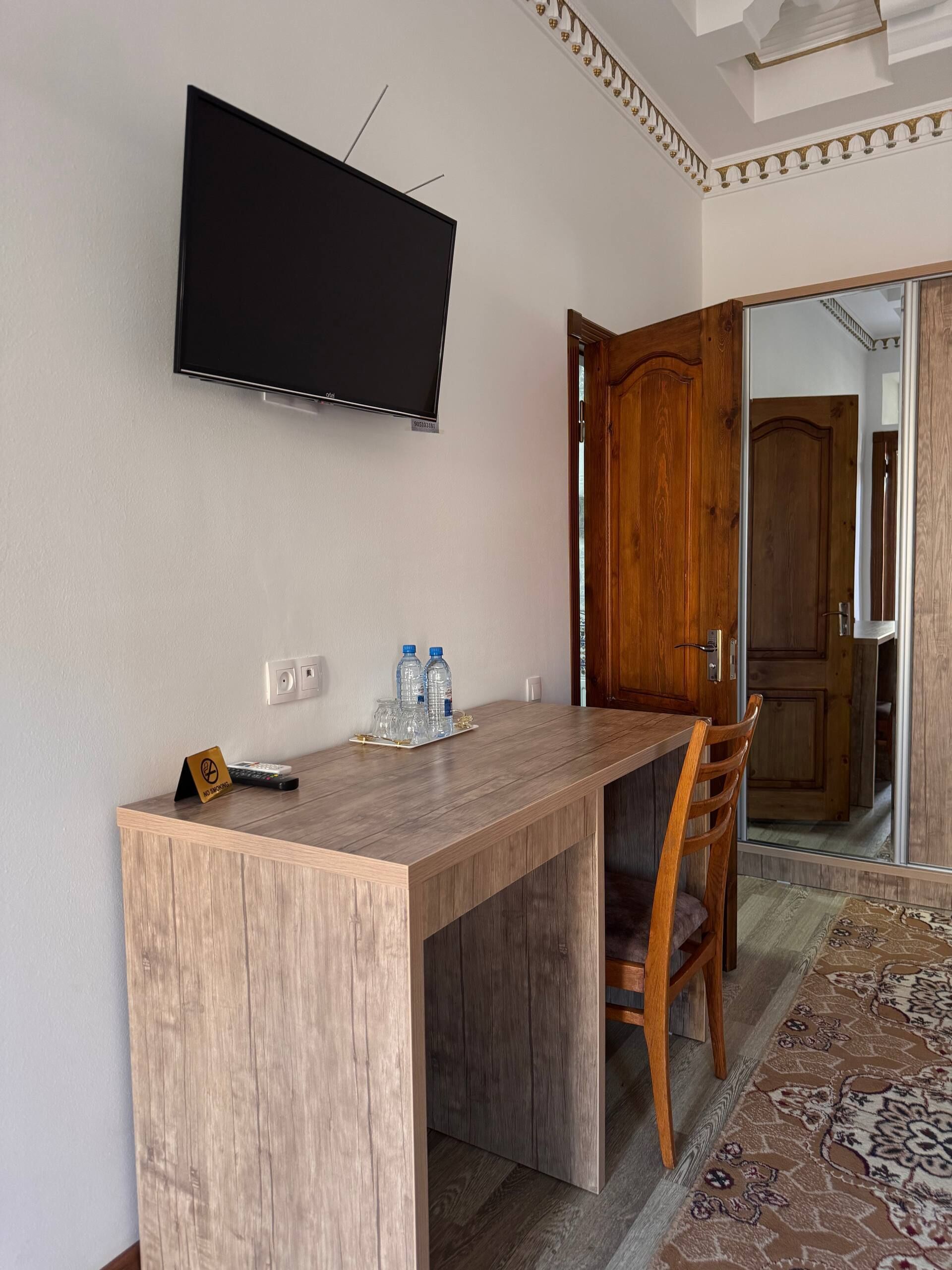 Quarto com escrivaninha de madeira, televisão fixada na parede, porta que dá para outro aposento e tapete estampado no chão.