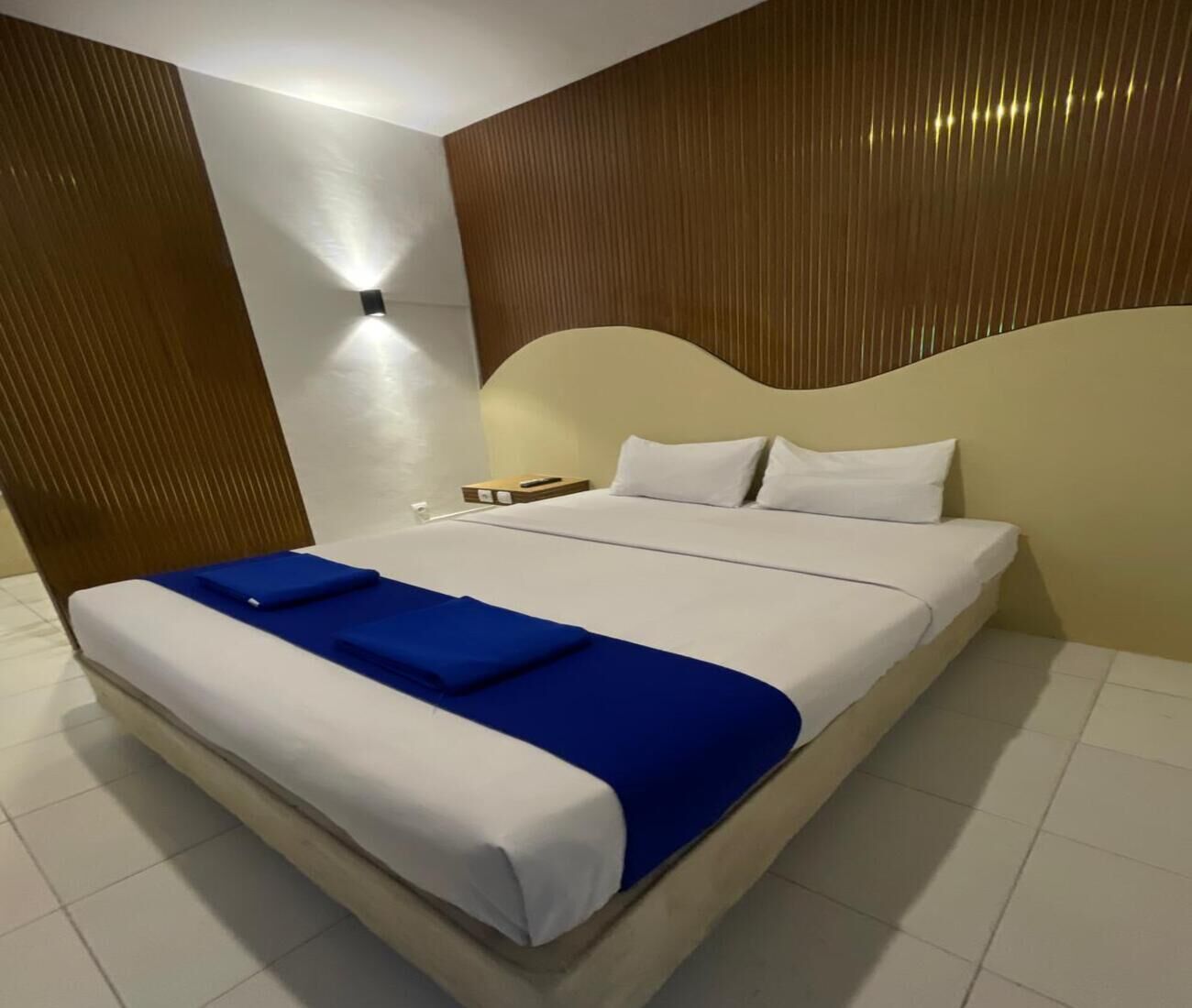Quarto de hotel moderno com uma cama grande, mesa de cabeceira e luminária fixada na parede.