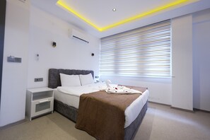 Quarto de hotel moderno com cama, mesas de cabeceira, janela com persianas e teto com iluminação embutida.