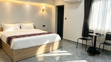 Quarto de hotel com cama, uma pequena mesa com cadeiras, luminária na parede e uma porta.