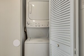 Máquina de lavar compacta e branca com painel de controle, instalada numa pequena lavanderia com porta e interruptor de luz na parede.