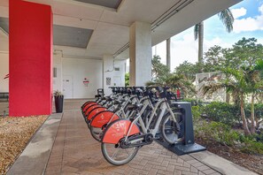Várias bicicletas estão estacionadas em frente a um edifício com um pilar vermelho e uma placa que indica um depósito para encomendas.