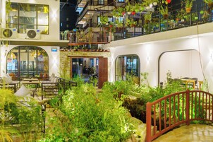 Um jardim interno com uma ponte de madeira, mesas e cadeiras, rodeado por diversas plantas e vegetação.