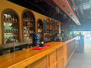 Uma área de bar com prateleiras cheias de garrafas de bebidas, copos e um balcão de madeira.