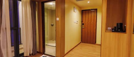 Quarto de hotel com piso de madeira, divisória de chuveiro em vidro, guarda-roupa de madeira e um tapete pequeno com chinelos.