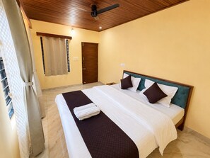 Quarto de hotel com cama, ventoinha no teto, janela com cortinas e porta.