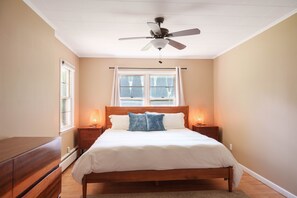 Quarto com cama de madeira, duas luminárias de apoio, ventilador de teto e janela com cortinas.