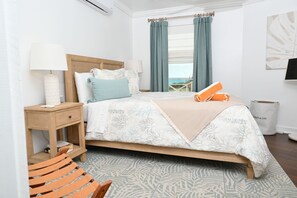 Quarto de hotel com cama, mesa de cabeceira, abajur, janela com cortinas e vista para o mar, além de uma toalha sobre a cama.