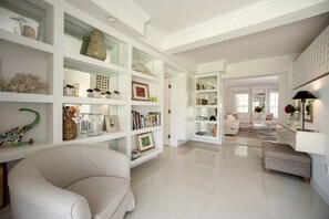 Ampla sala de estar com um sofá branco, uma mesa de centro e uma estante repleta de objetos decorativos.
