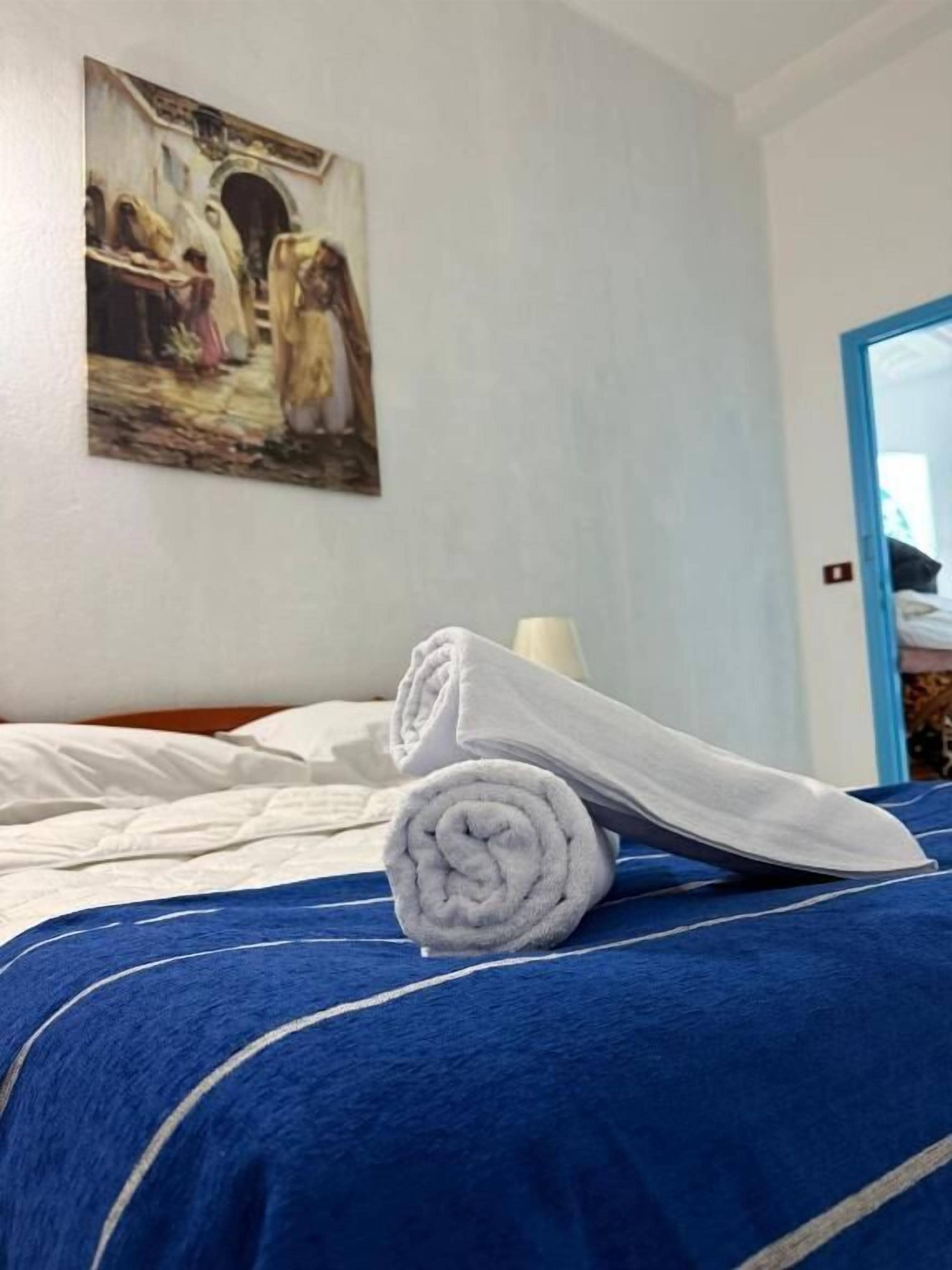 Cama bem arrumada com colcha azul e toalhas brancas dobradas, um quadro na parede e uma porta azul.