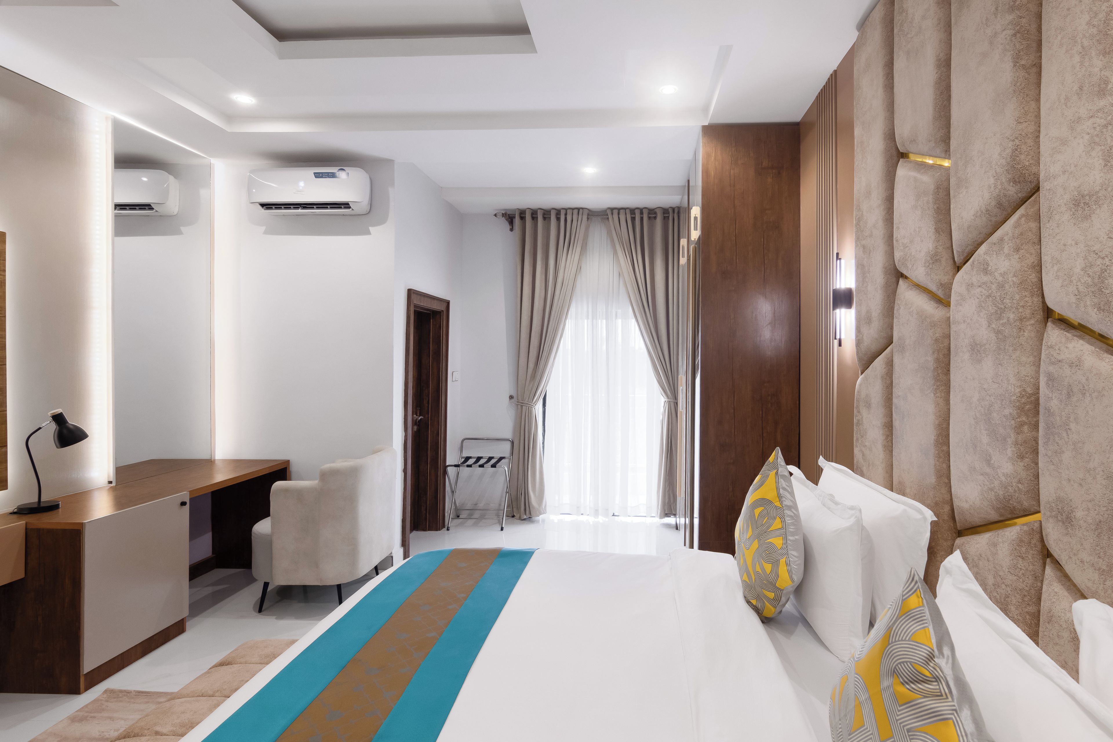 Quarto de hotel moderno com cama, mesa de cabeceira, abajur e vista para o corredor.