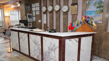 Balcão de recepção de hotel com bancada de mármore, vários relógios, certificados, bandeiras e uma pintura de uma cena de praia.