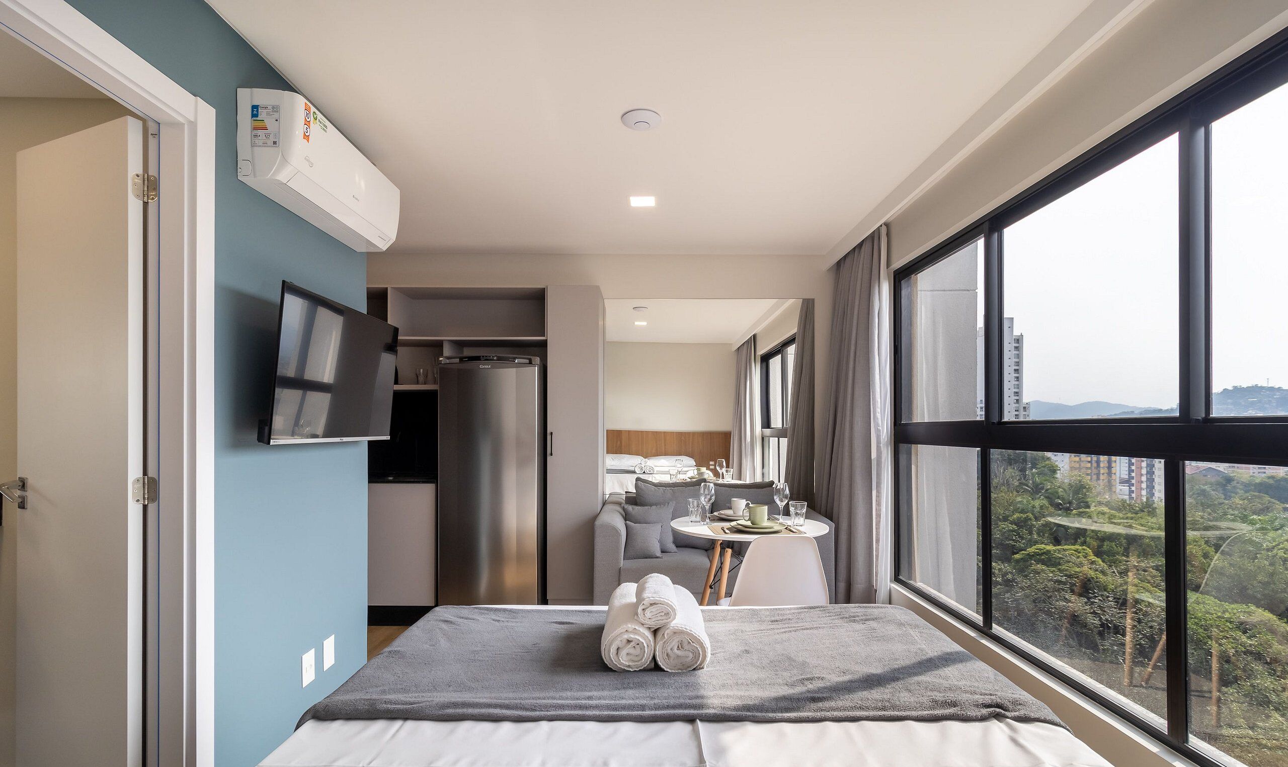 Quarto de hotel com cama, TV, minibar, área de refeições e uma grande janela com vista para a cidade.