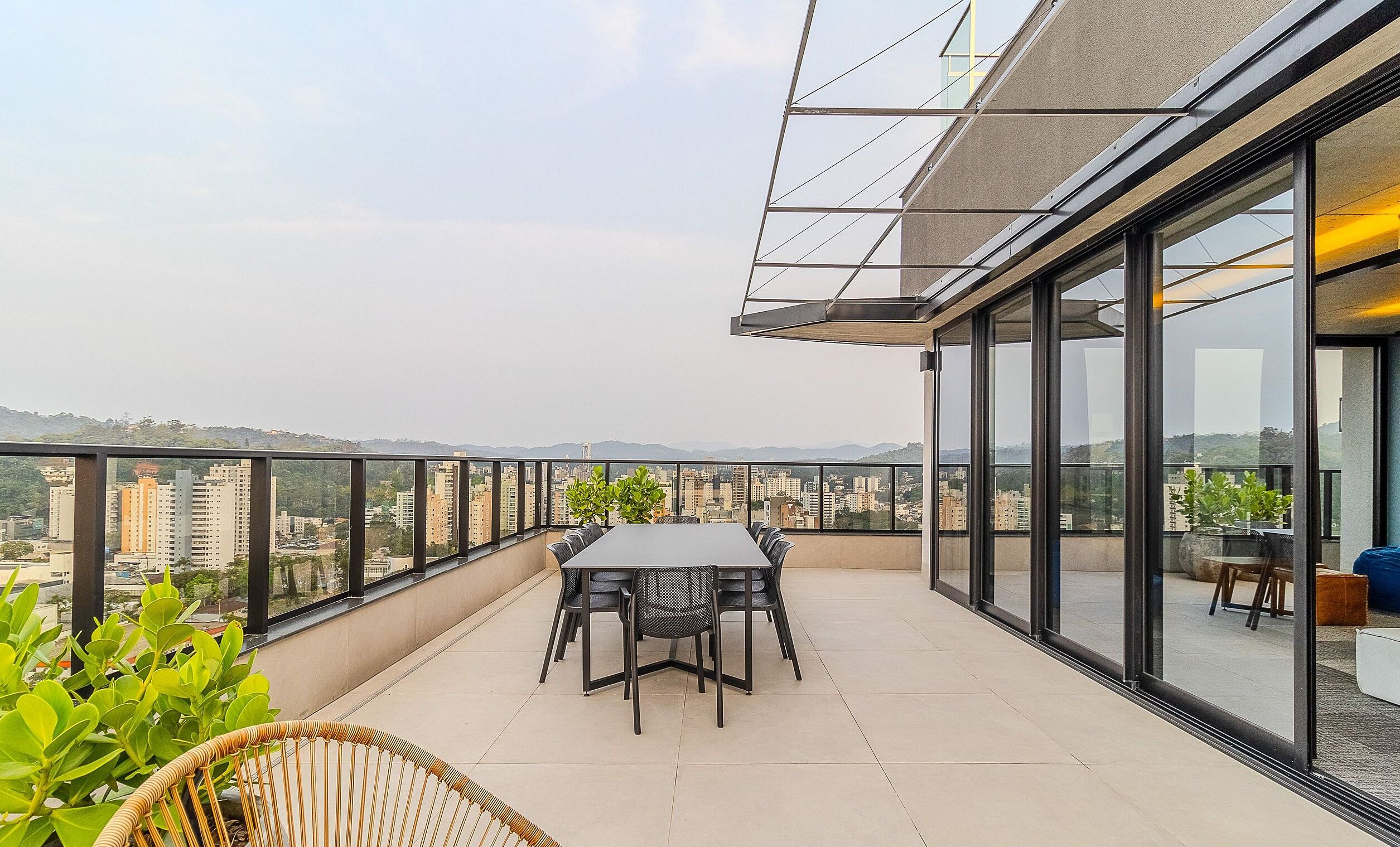 Terraço no telhado com mesa e cadeiras para refeições, cercado por paredes de vidro com vista para a cidade.