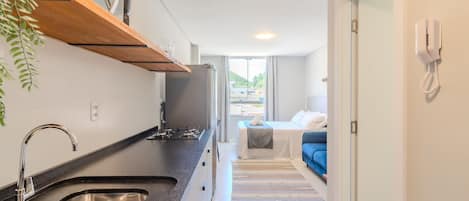 Quarto de hotel compacto com cozinha compacta, uma cama com cabeceira azul, mesa de cabeceira, telefone e vista para o exterior através de uma janela.