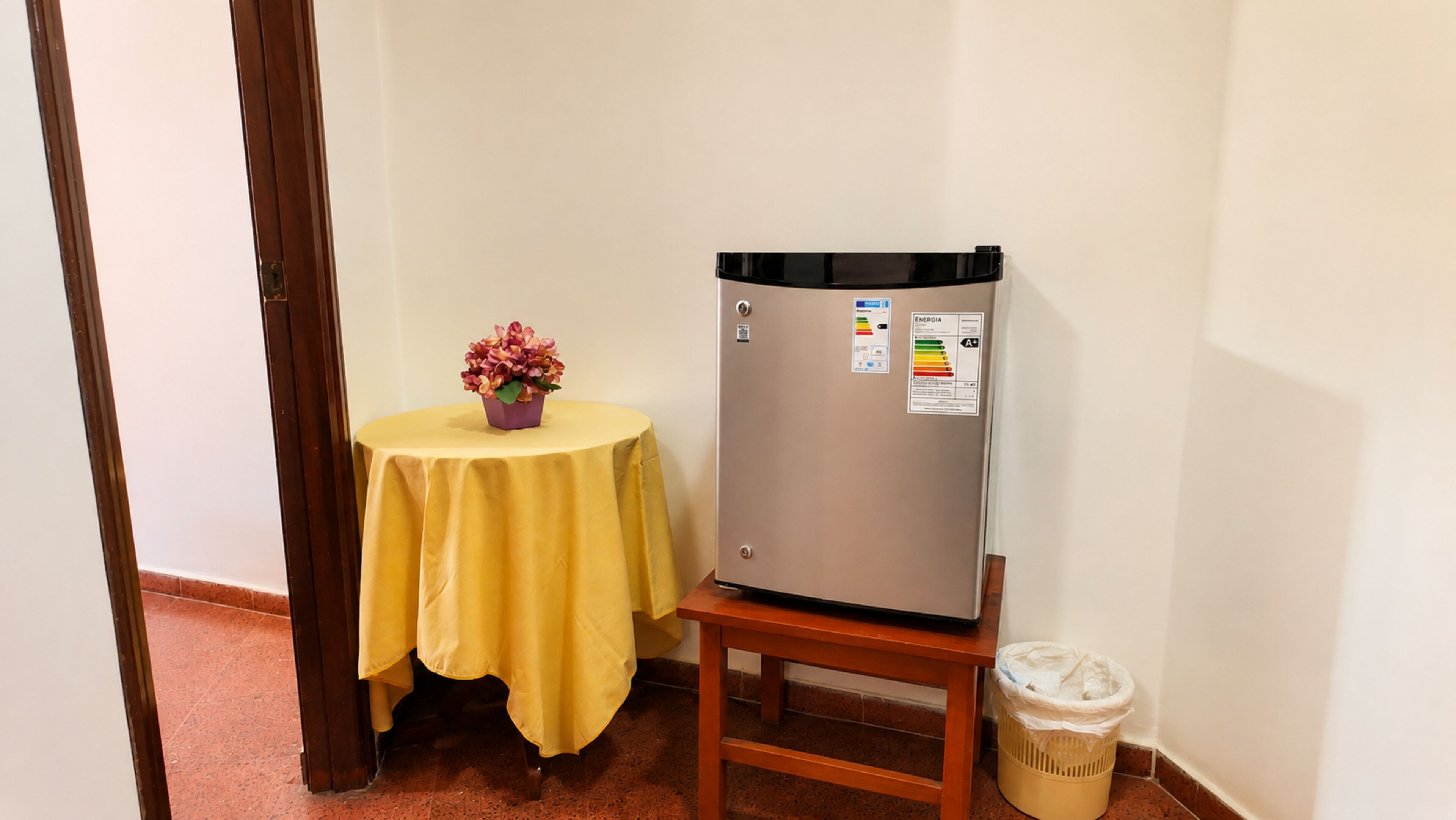 Quarto compacto com uma pequena mesa, geladeira e lixeira.