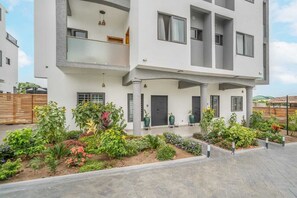 Um edifício residencial moderno de dois andares, com varanda, jardim frontal com diversas plantas e entrada com calçada.