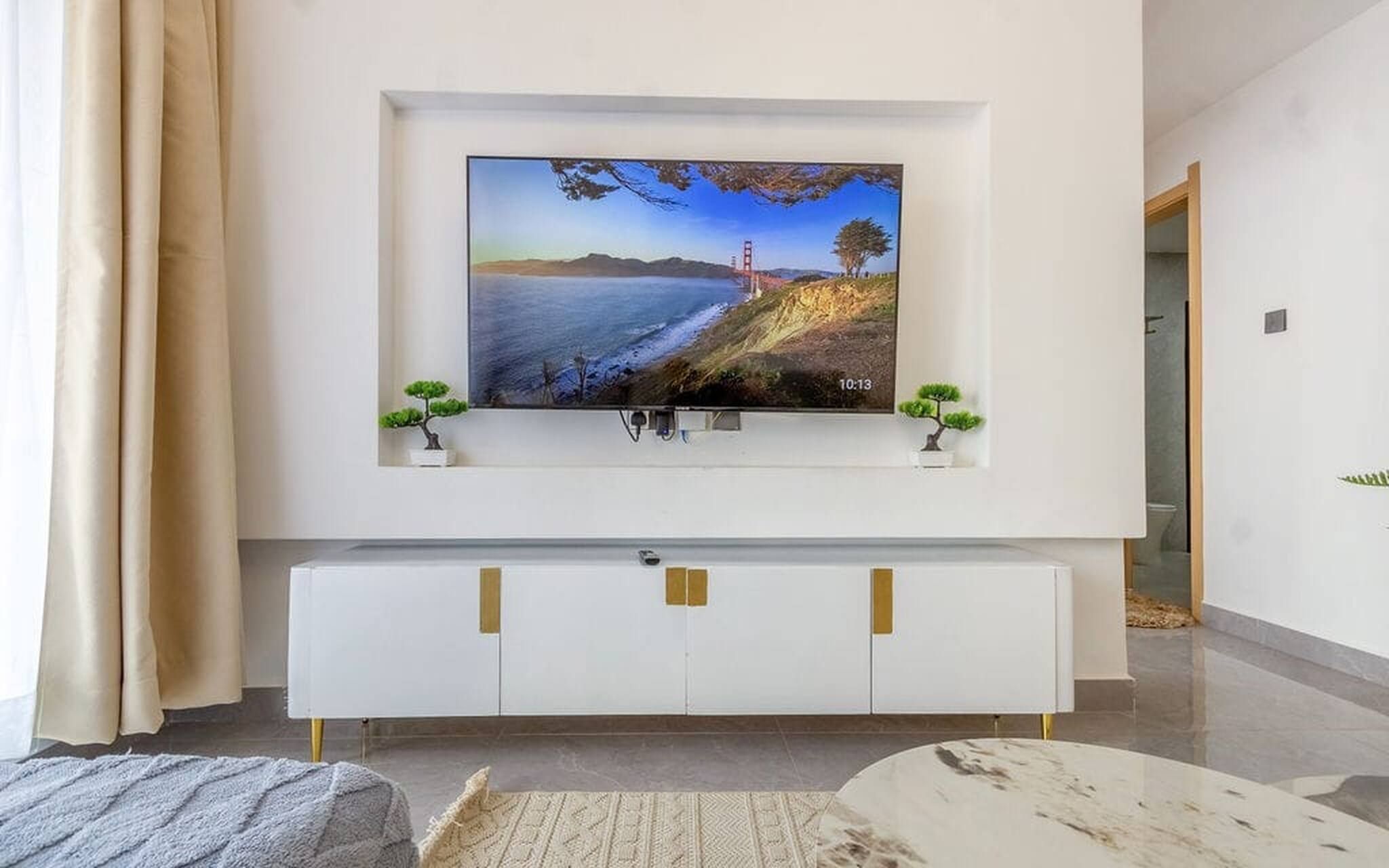 Uma sala de estar moderna com uma TV de tela plana presa a uma parede branca, um móvel branco embaixo e duas pequenas plantas em vasos de cada lado.