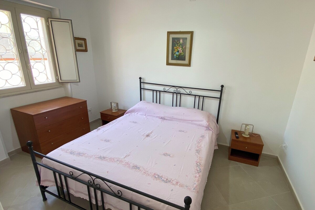 Quarto com cama, penteadeira, mesas de cabeceira e janela com detalhes decorativos.