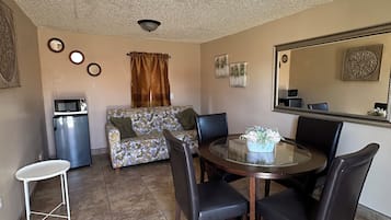 Uma sala de jantar com mesa redonda, cadeiras, ventilador de teto e área de cozinha com geladeira e micro-ondas.