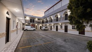 Pátio com chão pavimentado, uma van branca e um edifício com várias varandas e portas.