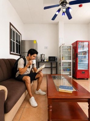 Uma pessoa sentada em um sofá em um quarto com ventilador de teto, geladeira, dispensador de bebidas vermelho e mesa de centro de madeira.