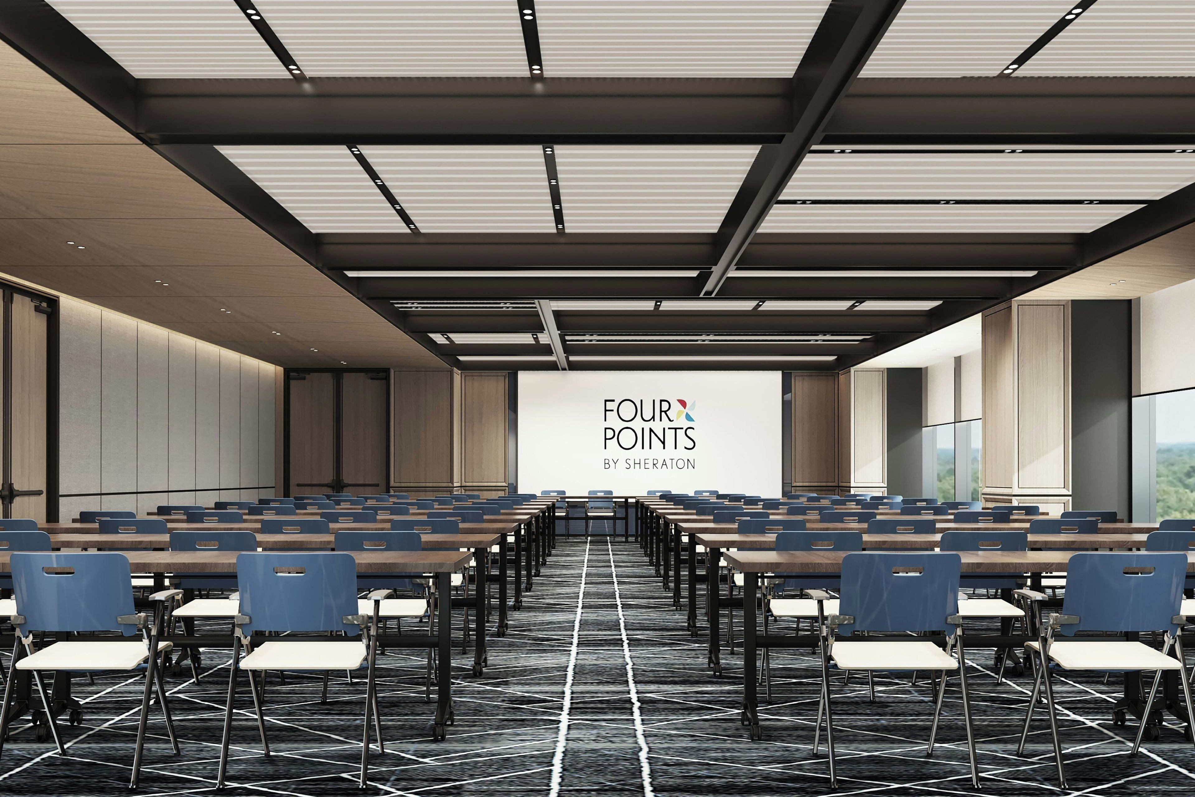 Uma sala de conferências com fileiras de mesas e cadeiras, uma tela grande exibindo 'FOUR POINTS BY SHERATON' e vista para o exterior através de amplas janelas.