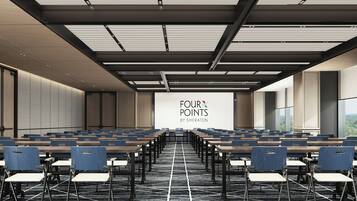 Uma sala de conferências com fileiras de mesas e cadeiras, uma tela grande exibindo 'FOUR POINTS BY SHERATON' e vista para o exterior através de amplas janelas.