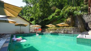 Piscina com piso azulejado, cercada por espreguiçadeiras e guarda-sóis, com uma parede de pedra e vegetação ao fundo.