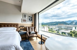Quarto de hotel com cama, cadeira, escrivaninha e uma grande janela com vista para a paisagem urbana e montanhas.