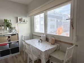 Uma área de refeições com mesa e cadeiras, uma janela com vista para prédios e armários com contentores.