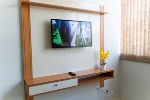 Uma televisão fixada na parede com moldura, um controle remoto e uma pequena prateleira com um vaso de flores.
