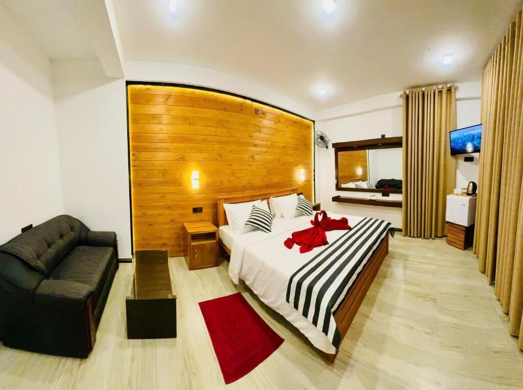 Quarto de hotel moderno com cabeceira de madeira, cama com roupa de cama listrada, manta vermelha, sofá preto e televisão de tela plana.
