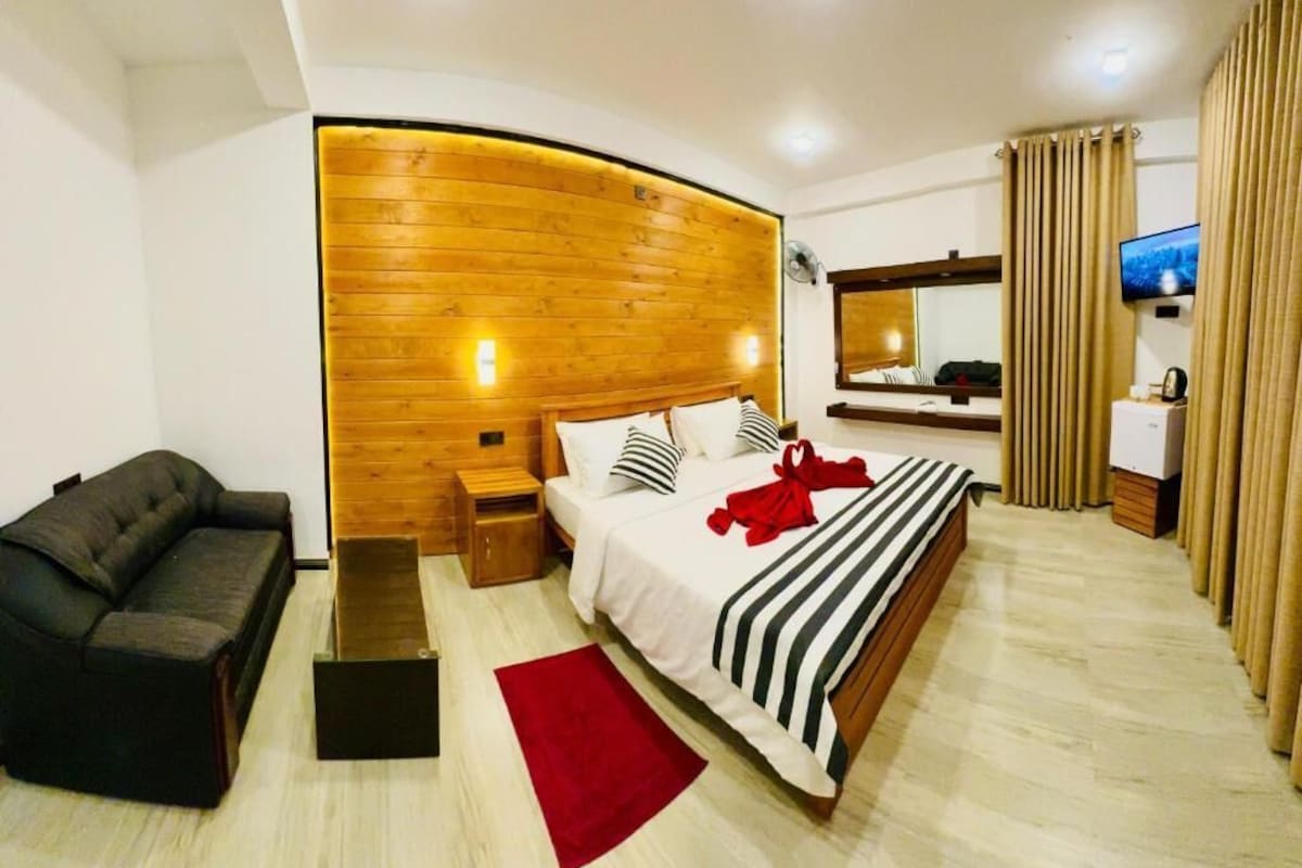 Quarto de hotel moderno com cabeceira de madeira, cama com roupa de cama listrada, manta vermelha, sofá preto e televisão de tela plana.