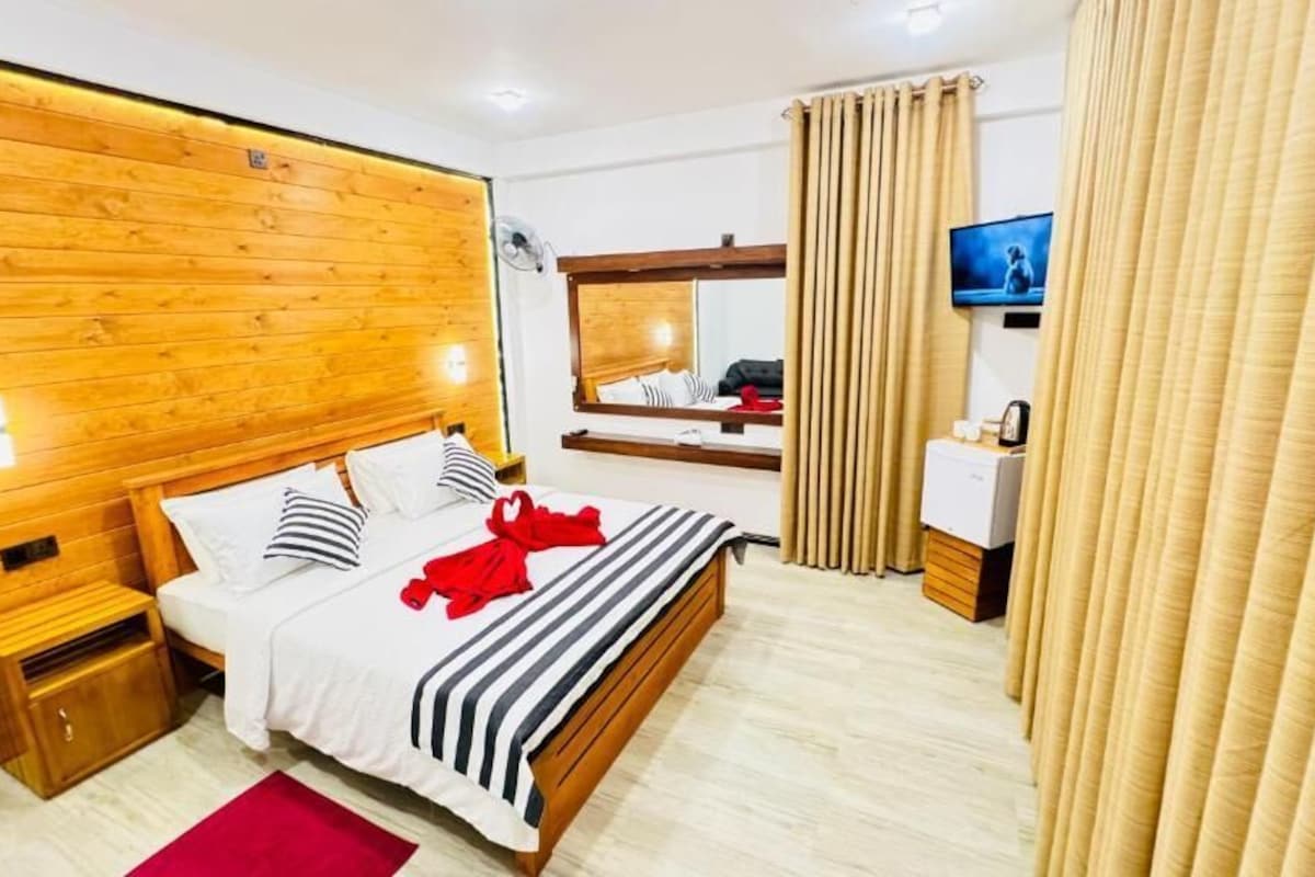 Quarto de hotel com cabeceira de madeira, cama com roupa de cama listrada, toalha vermelha, televisão na parede e um espelho.