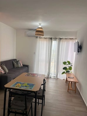 Uma sala de estar com sofá, mesa de jantar com cadeiras, luminária pendente, planta e televisão na parede.