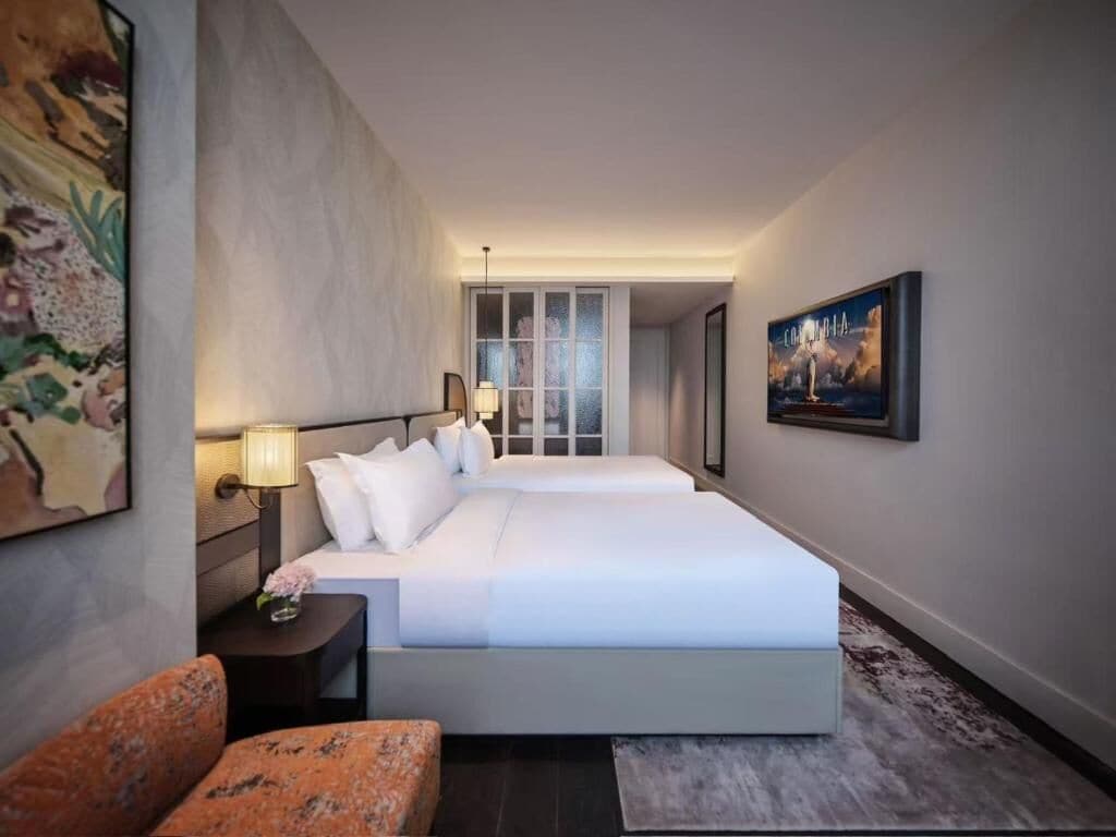 Quarto de hotel com cama grande, mesa de cabeceira, luminária, pintura e vista para o exterior.
