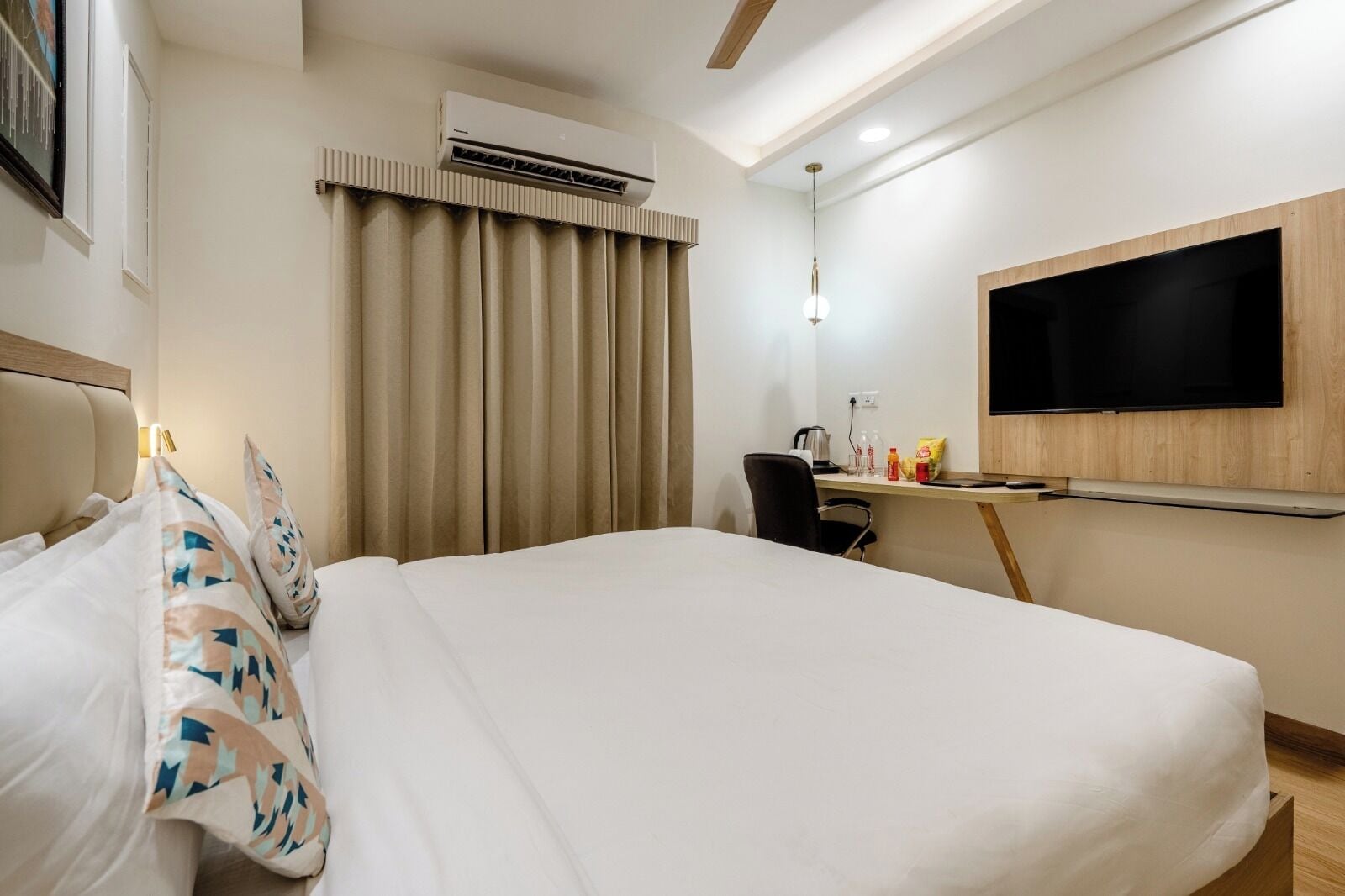 Quarto de hotel com cama, escrivaninha, cadeira, televisão, aparelho de ar condicionado e janela com cortinas.