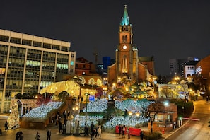 Uma igreja com torre do relógio, rodeada por árvores iluminadas e um edifício moderno ao fundo.