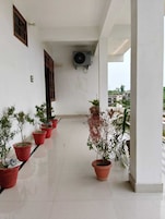 Corredor com plantas em vasos, um ventilador de teto e vista dos edifícios ao redor.